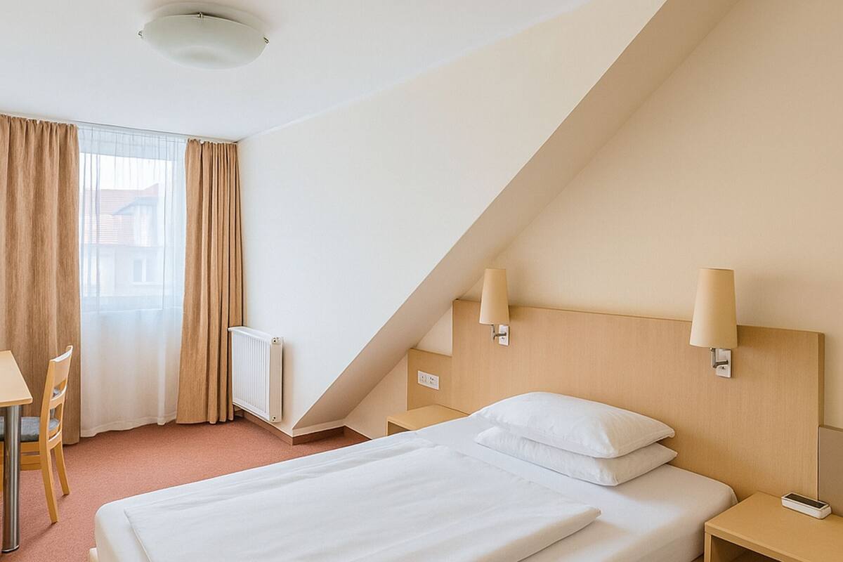 Quarto de hotel com uma cama de solteiro, uma secretária com cadeira, um radiador e duas luminárias fixadas na parede.