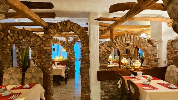 Uma área de jantar rústica com arcos de pedra, vigas de madeira e piso de azulejos. As mesas estão dispostas com toalhas brancas e guardanapos vermelhos.