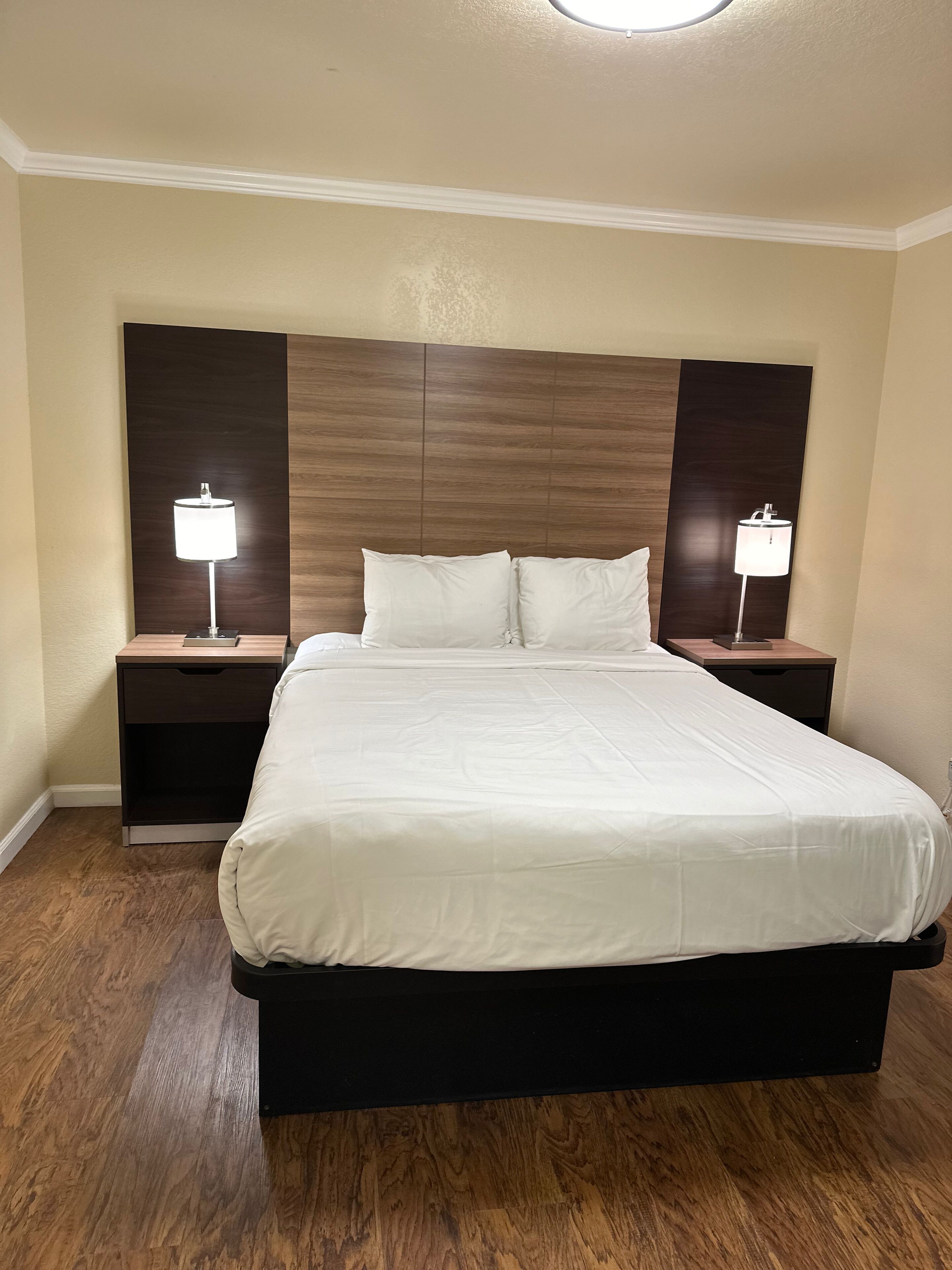 Cama bem arrumada com lençóis brancos, duas mesas de cabeceira com luminárias e um painel de cabeceira em madeira escura.