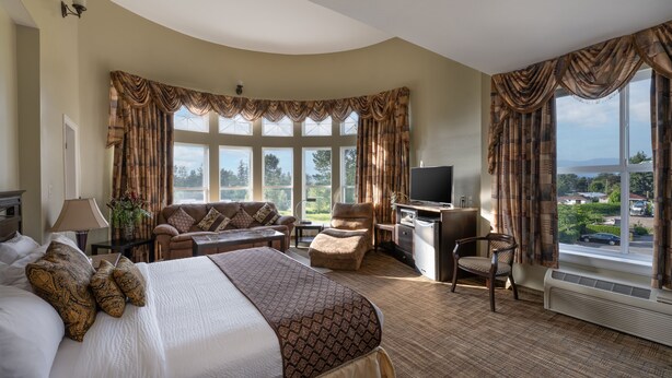 Quarto de hotel com uma cama grande, um sofá, uma escrivaninha e uma televisão. Há janelas amplas com cortinas e vista para o exterior.