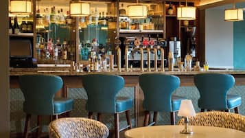 Uma área de bar com diversas opções de assento, balcão com vários tipos de bebidas e uma máquina de café, além de uma televisão.