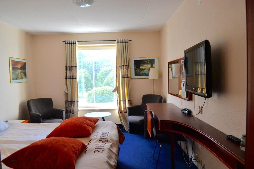 Quarto de hotel com uma cama, duas cadeiras, uma escrivaninha, televisão e janela com cortinas.