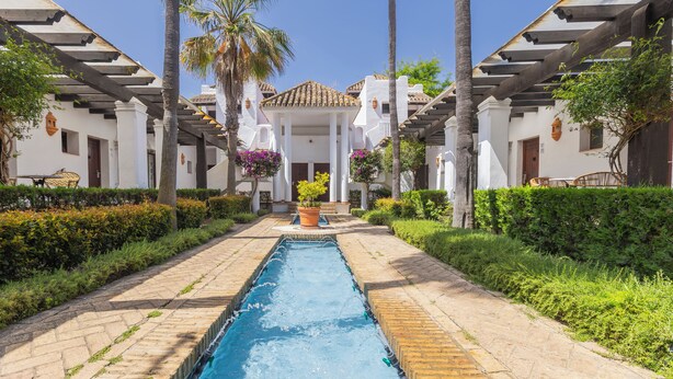 Piscina com borda em azulejo, rodeada por um jardim com palmeiras e um edifício branco com colunas.