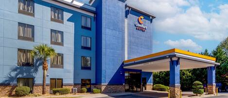 Um hotel com fachada azul, entrada coberta e placa visível da 'Comfort Suites'.