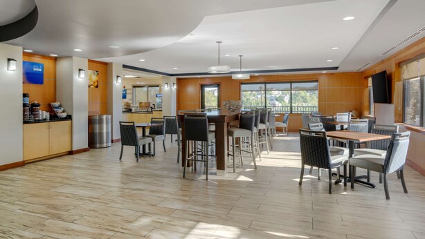 Uma sala de jantar ampla com várias mesas e cadeiras, área de cozinha com cafeteira, e grandes janelas que proporcionam luz natural.