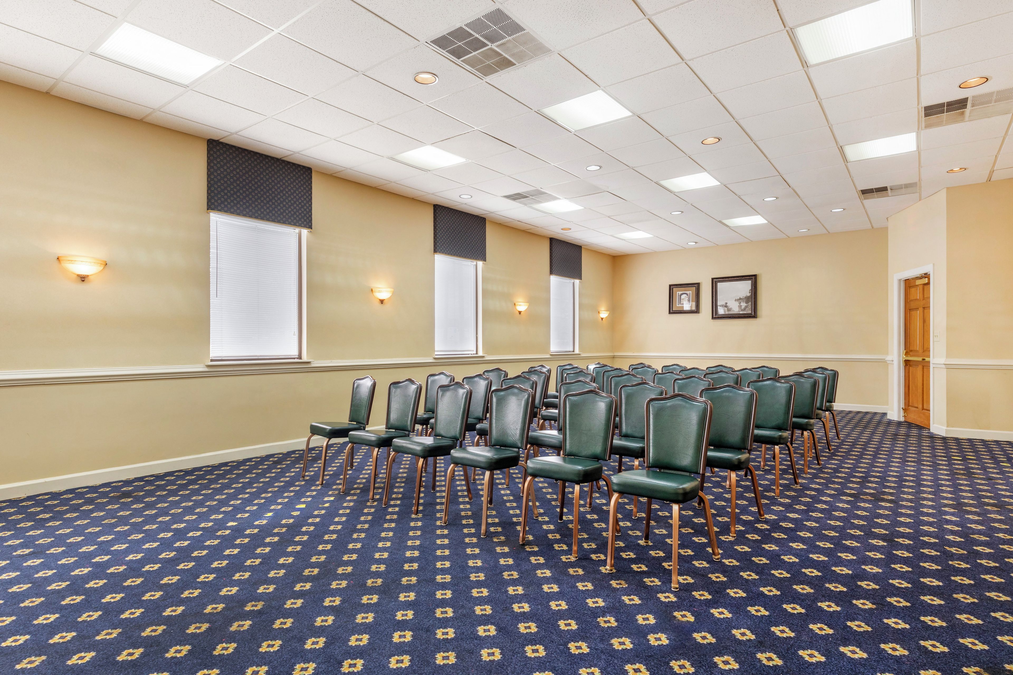 Sala de conferências com fileiras de cadeiras verdes, paredes bege e carpete estampado.