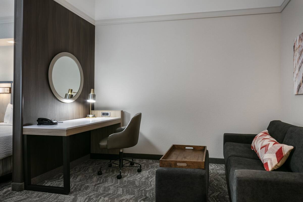 Quarto de hotel com cama, escrivaninha com cadeira, mesa pequena, sofá e espelho fixado na parede.
