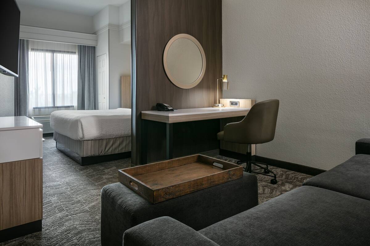 Quarto de hotel com uma cama, uma escrivaninha com cadeira, mesa de cabeceira, abajur e bandeja de madeira.