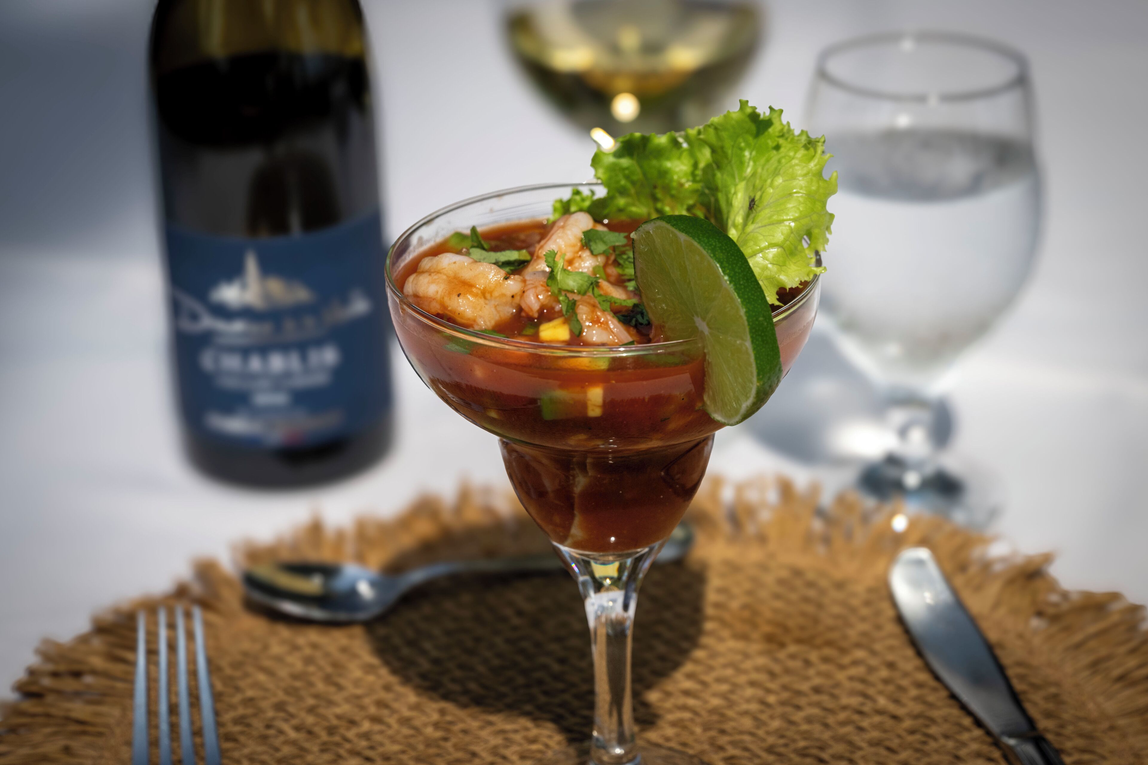 Um cocktail num copo com camarão, lima e alface, uma garrafa de vinho e um copo de água sobre uma mesa com garfo e faca.