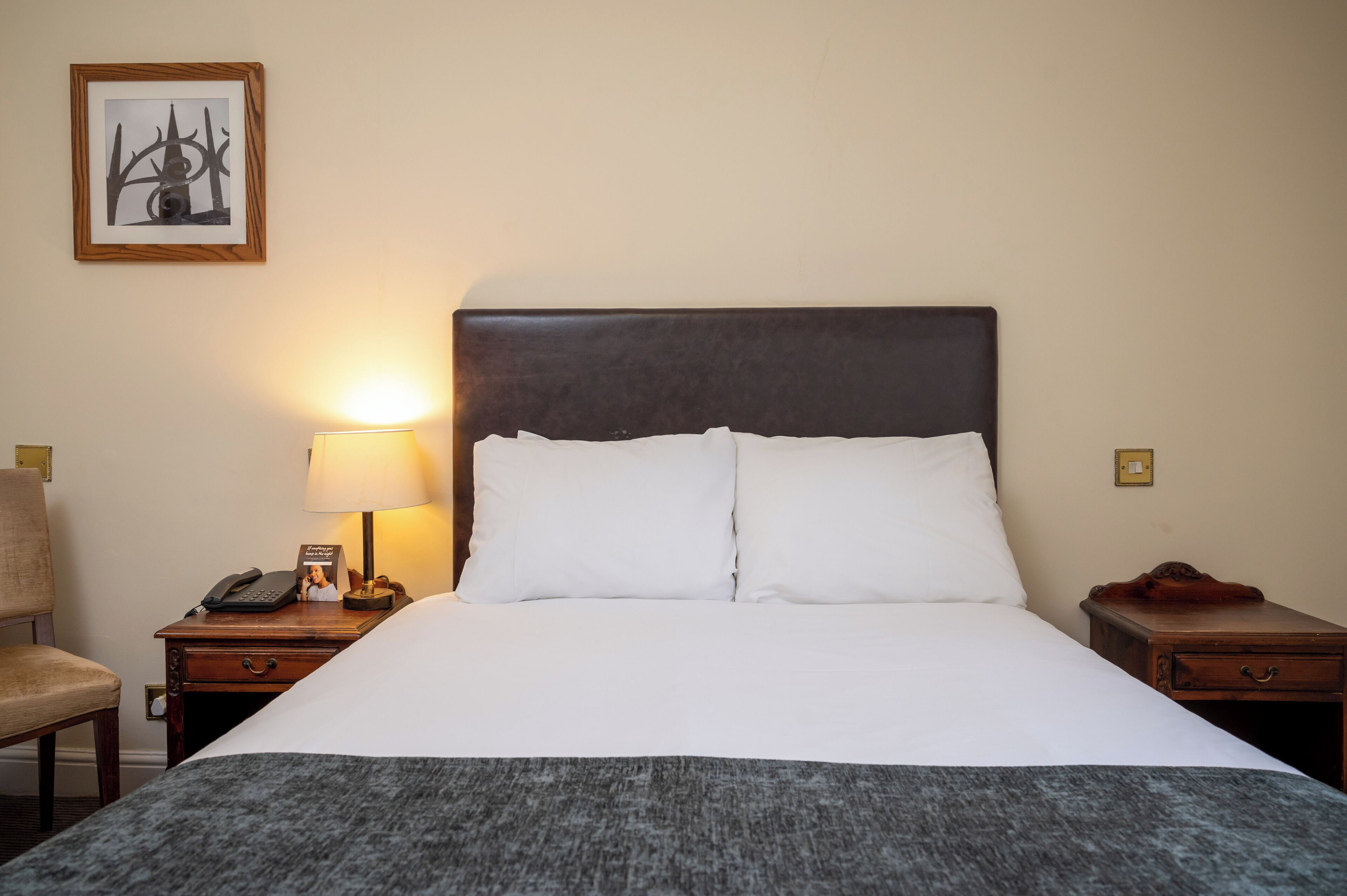 Quarto de hotel com uma cama grande, abajur, telefone, quadro na parede e mesa de cabeceira de madeira.