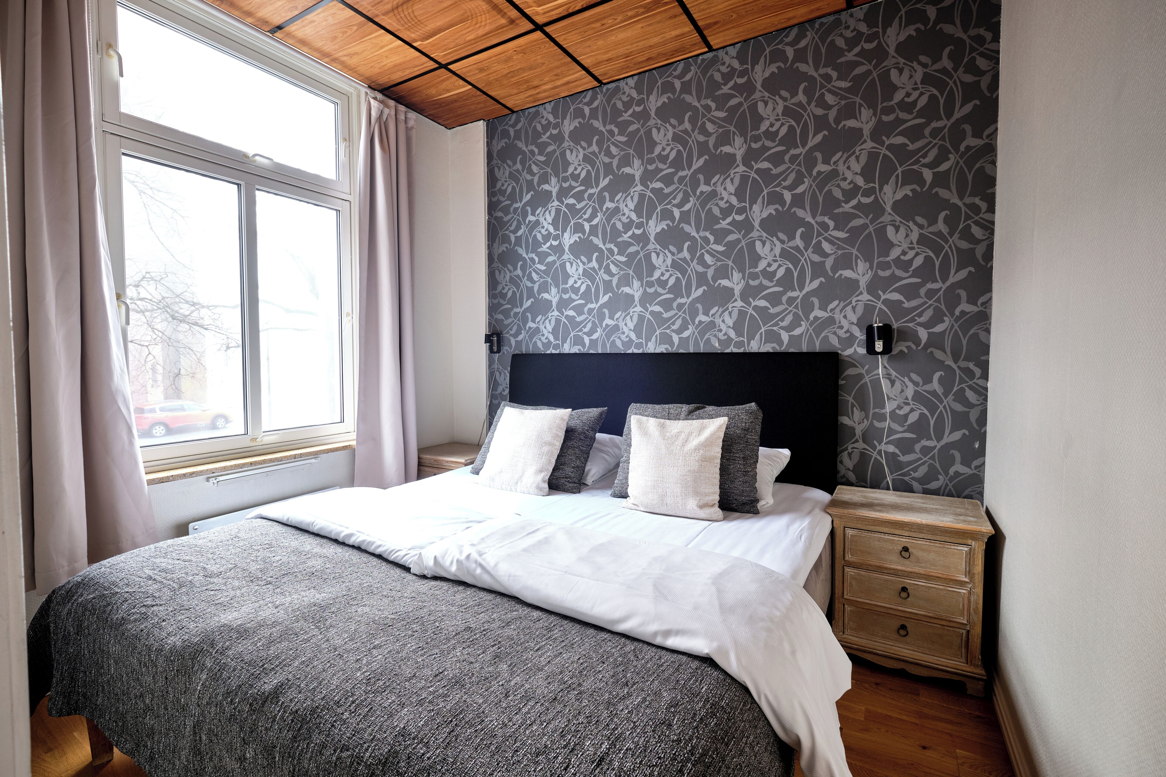 Quarto com cama, mesa de cabeceira, janela com cortinas e parede estampada.