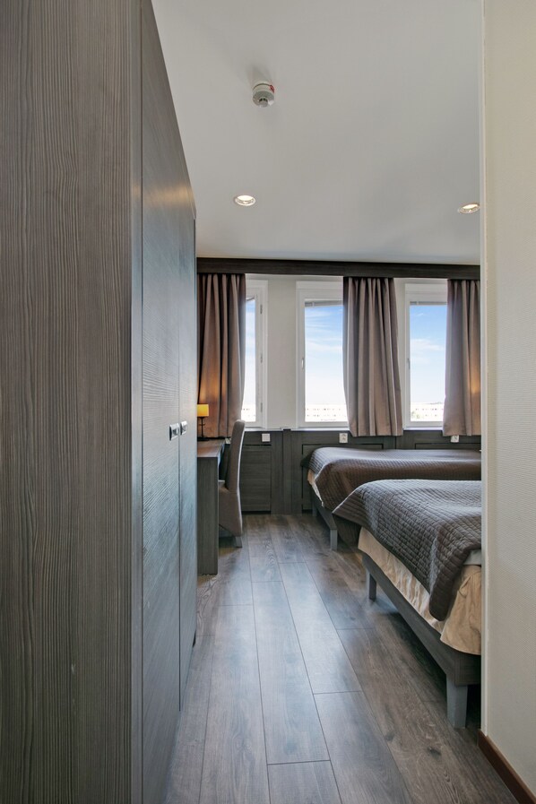 Quarto de hotel estreito com piso de madeira, duas camas, uma escrivaninha e uma janela com cortinas.