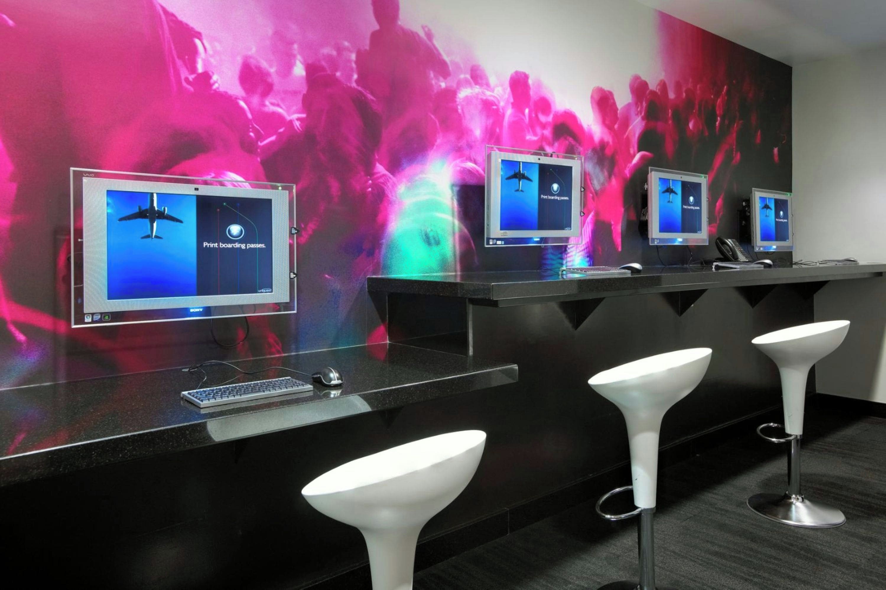 Uma área de bar moderna com três banquetas, um balcão com monitor de computador e um vibrante mural abstrato na parede.