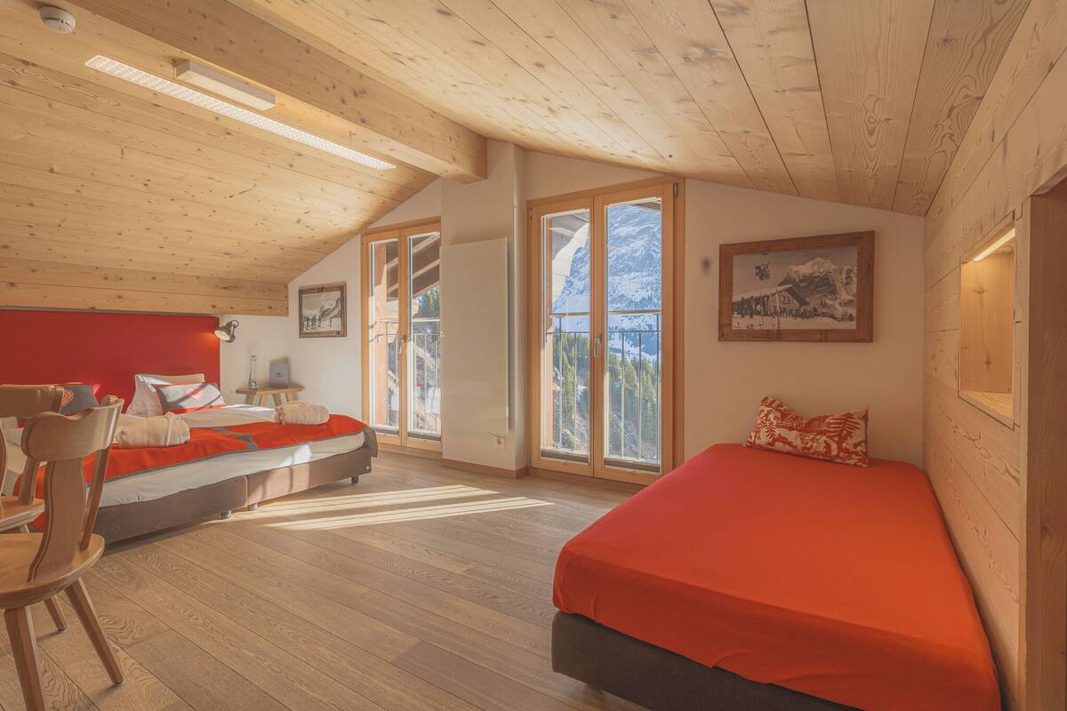 Quarto moderno com teto de madeira, cama com roupa de cama vermelha, escrivaninha com cadeira e amplas janelas com vista para as montanhas.