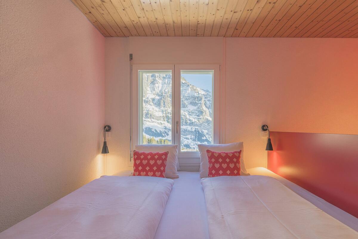 Quarto com duas camas, janela com vista para as montanhas e teto de madeira.