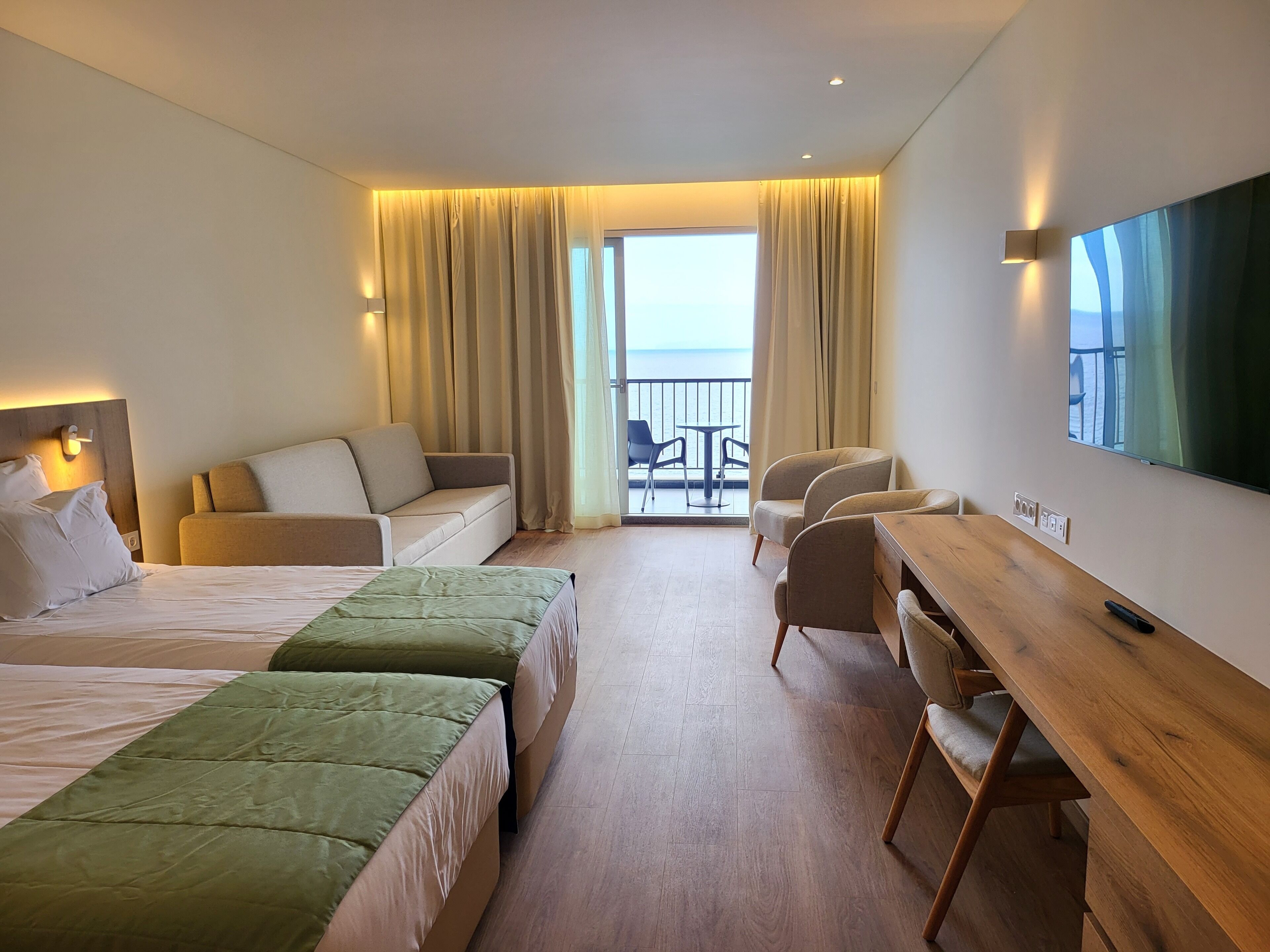 Quarto de hotel com duas camas, um sofá, uma secretária de madeira e uma televisão.