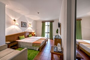 Quarto de hotel com duas camas, um sofá, uma mesinha com uma chaleira e uma janela com cortinas verdes.