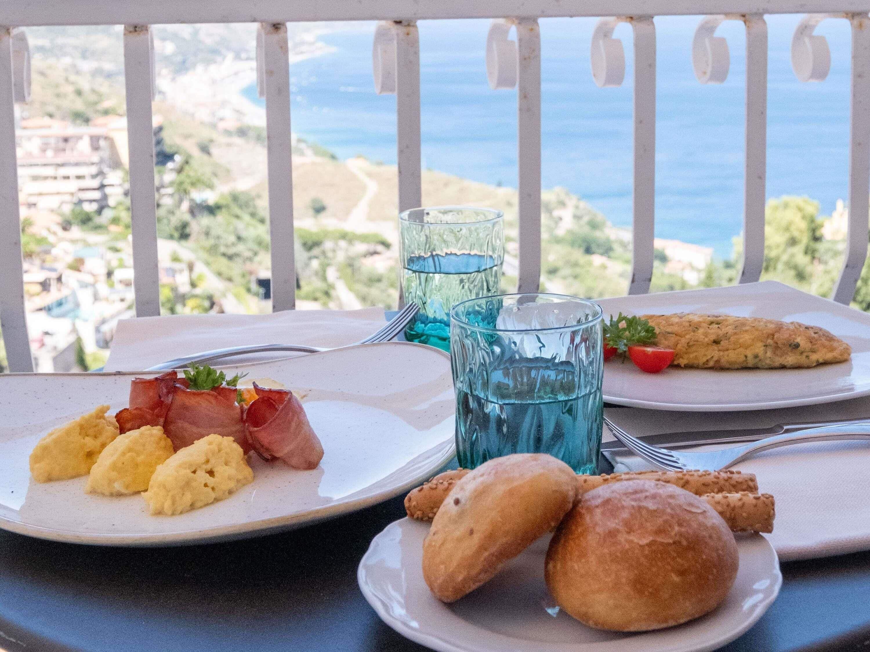 Mesa com itens de café da manhã, como ovos, bacon e pão, com vista panorâmica do mar e edifícios ao fundo.