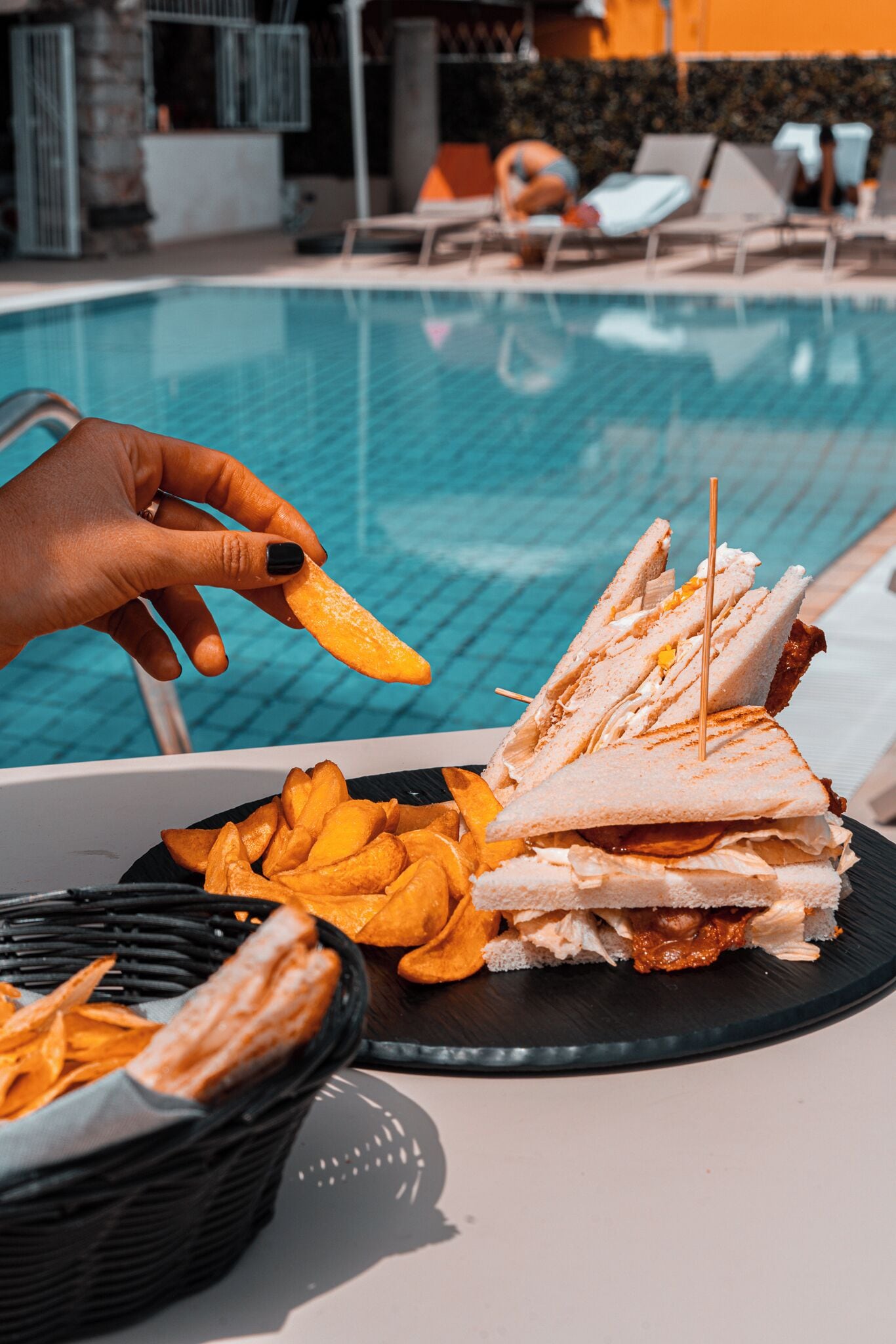 Disposição para refeições à beira da piscina, com sanduíche e batatas fritas, uma mão segurando uma batata frita e cadeiras de lounge ao fundo.