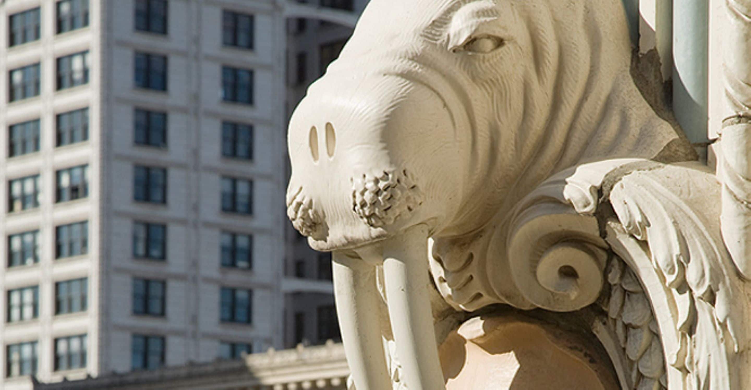 Close-up de uma escultura arquitetônica branca com detalhes intrincados, com um edifício moderno e várias janelas ao fundo.