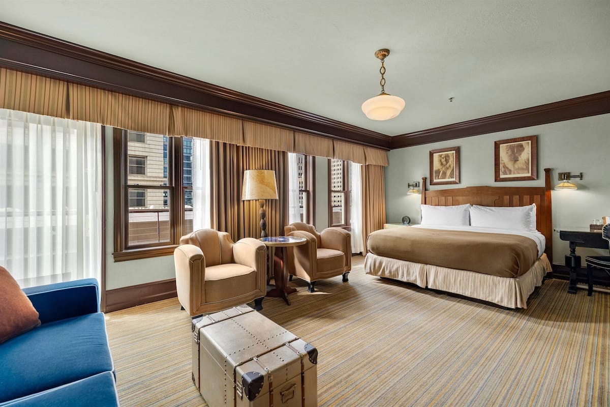 Quarto de hotel com uma cama grande, duas poltronas, uma mesinha, uma mala e vista da cidade através de grandes janelas.
