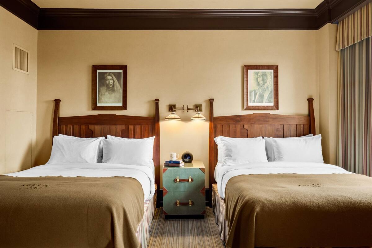 Quarto de hotel com duas camas, uma mesa de cabeceira com abajur e quadros nas paredes.