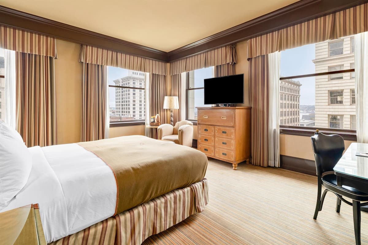 Quarto de hotel com uma cama grande, televisão de tela plana, cômoda, uma cadeira e vista para edifícios da cidade.