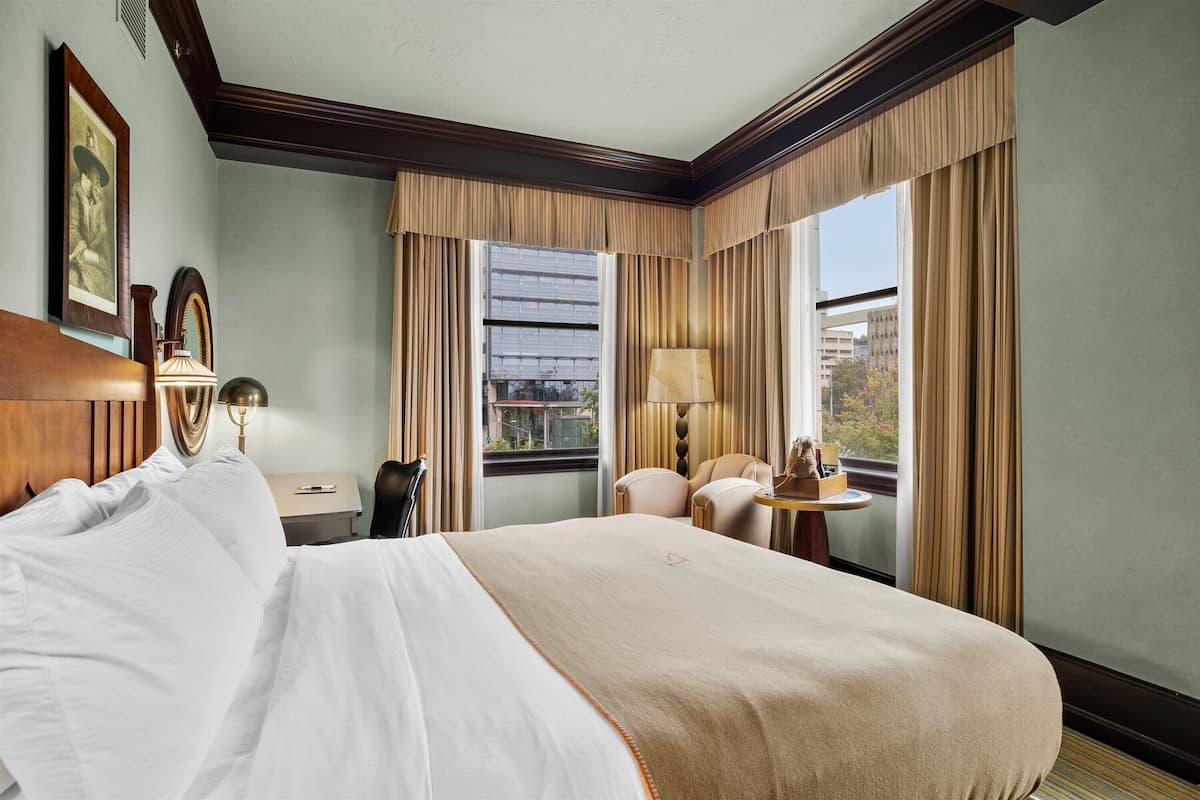 Quarto de hotel com cama grande, escrivaninha, cadeira e janela com cortinas.