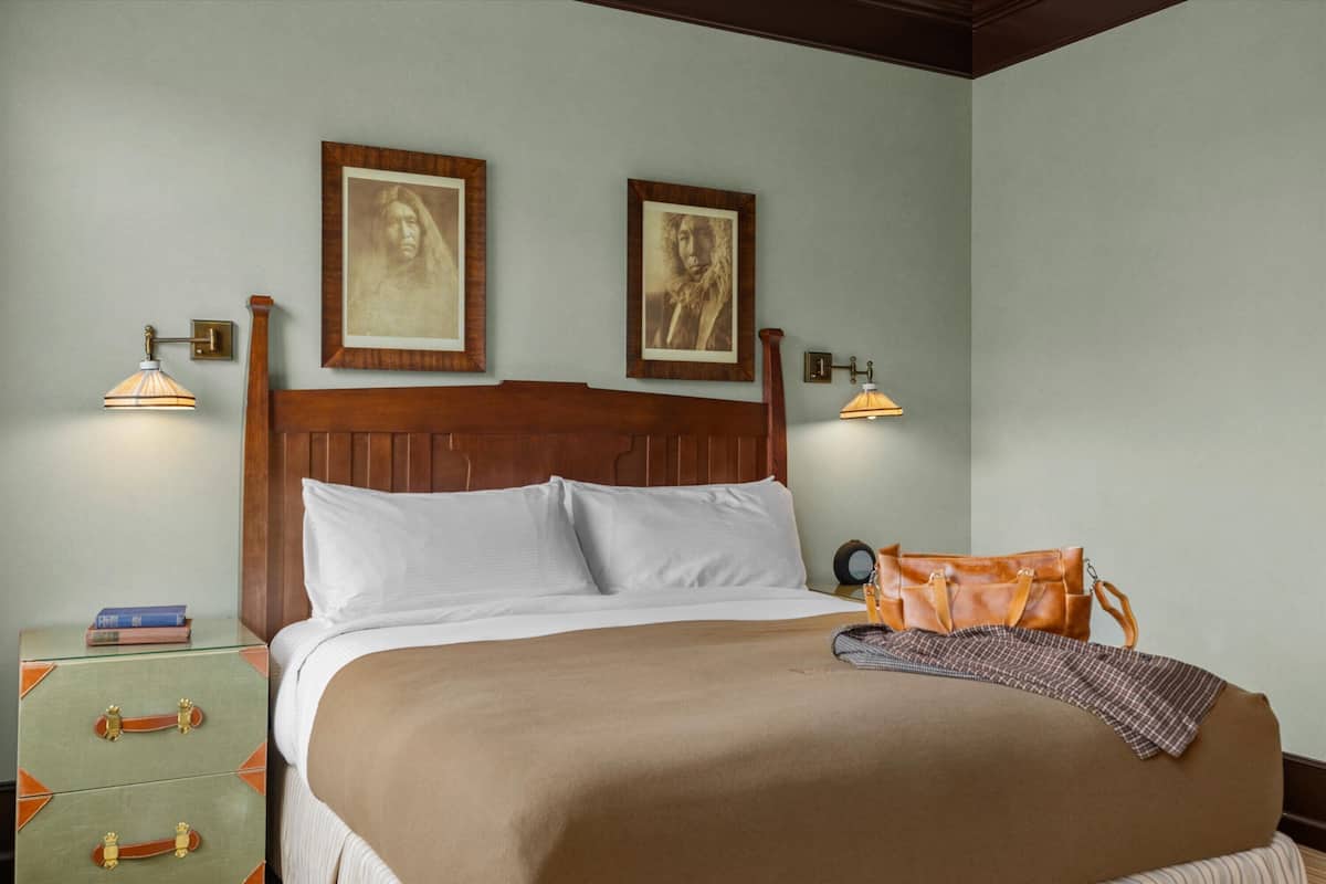Uma cama bem arrumada com cabeceira de madeira, duas luminárias na parede e uma pequena mesa de cabeceira com um livro e uma bolsa.
