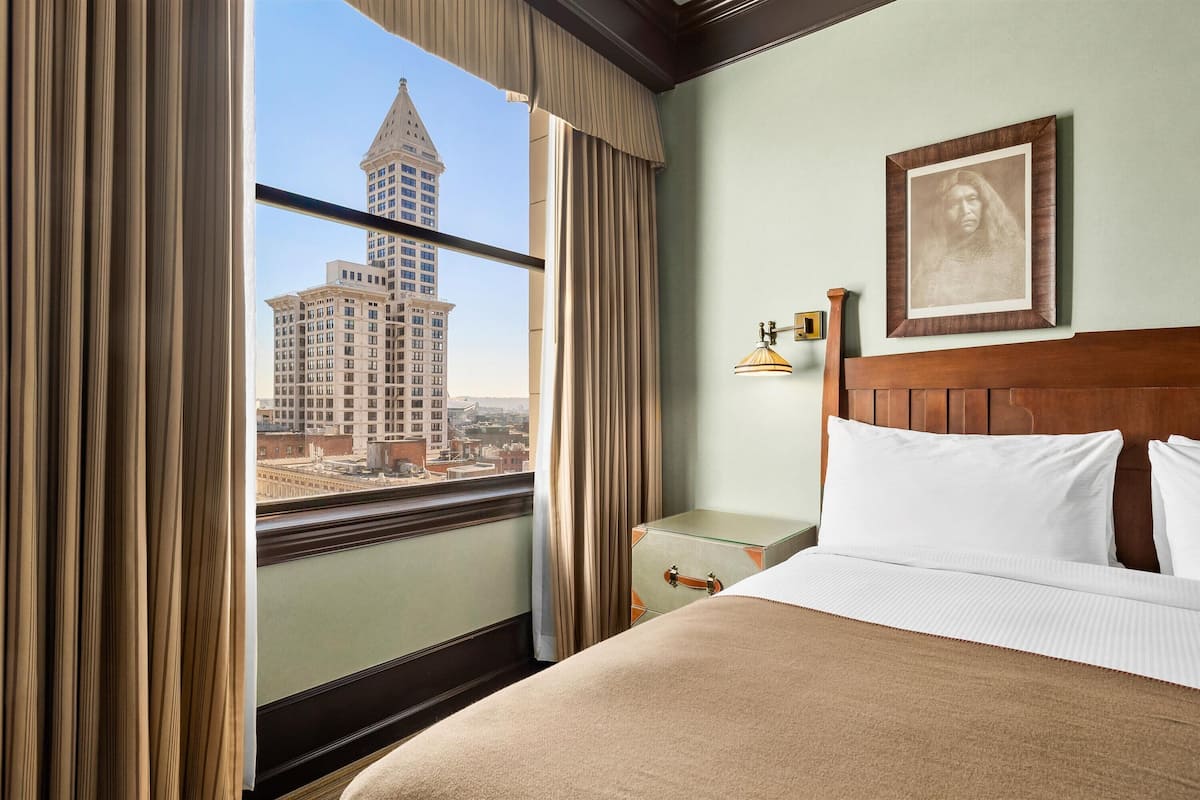 Quarto de hotel com uma grande janela, uma cama, mesa de cabeceira e um quadro na parede.