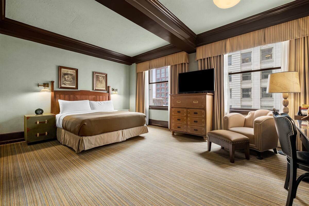 Quarto de hotel com uma cama grande, cômoda, televisão, cadeira e escrivaninha.