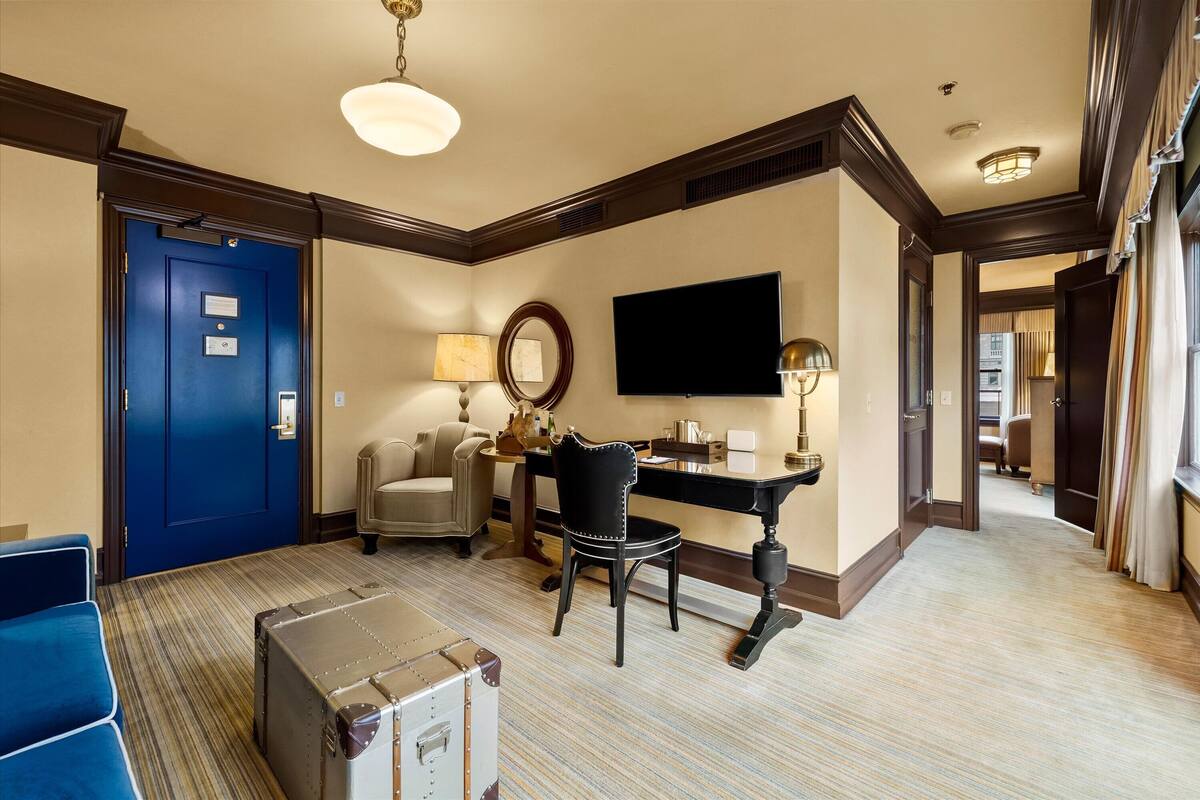 Quarto de hotel com porta azul, uma escrivaninha com luminária, uma cadeira, televisão e mala no chão.