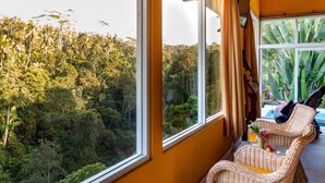 Quarto com grandes janelas que oferecem vista para uma floresta densa. Há uma cadeira de vime com almofada e uma mesa pequena com um vaso e flores.