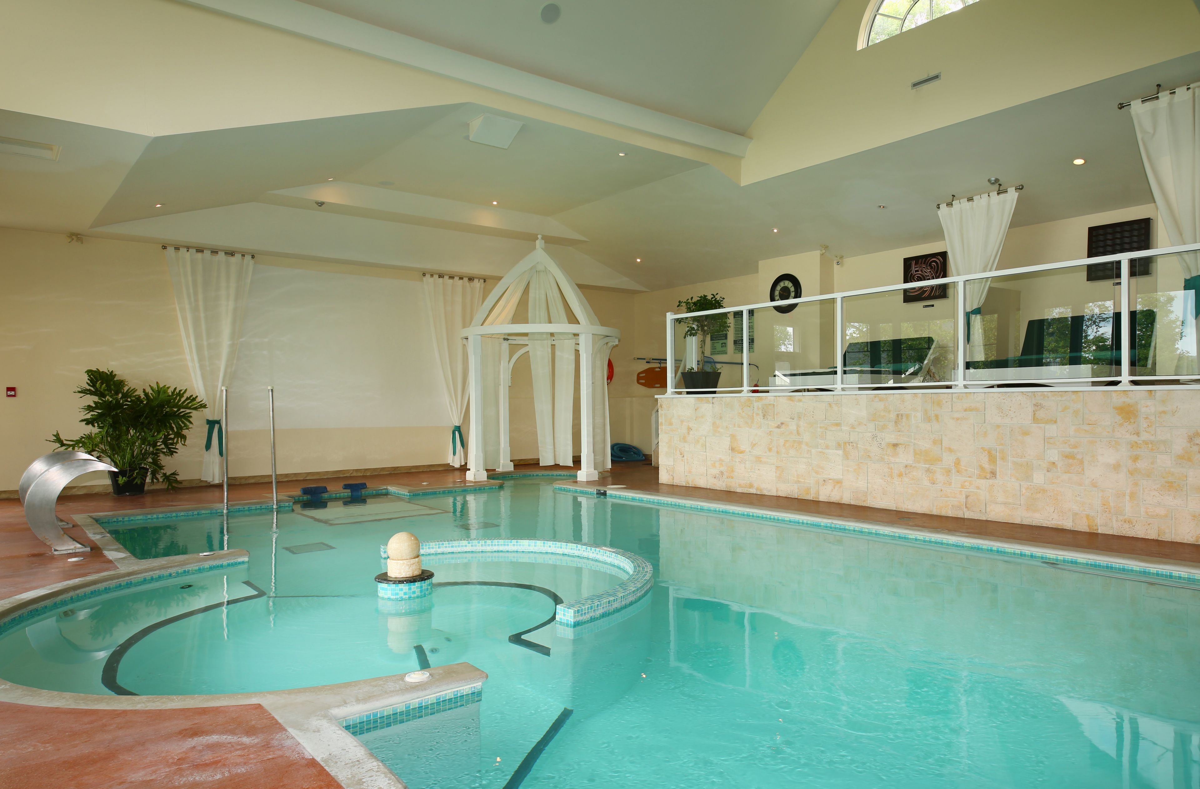 Piscina interna com um recurso aquático transparente, uma pequena ilha com uma esfera de água, e uma área de estar com paredes de vidro e vista para a piscina.
