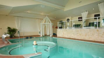 Piscina interna com um recurso aquático transparente, uma pequena ilha com uma esfera de água, e uma área de estar com paredes de vidro e vista para a piscina.