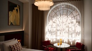 Quarto de hotel com cama, mesa redonda com cadeiras, um lustre e uma grande janela com cortinas.