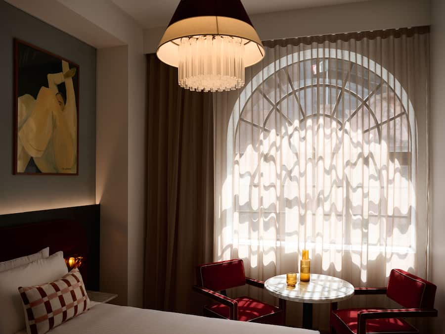 Quarto de hotel com cama, mesa redonda com cadeiras, um lustre e uma grande janela com cortinas.