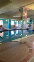 Piscina interior com água cristalina, uma cadeira de salva-vidas e área na beira da piscina com deck azulejado.
