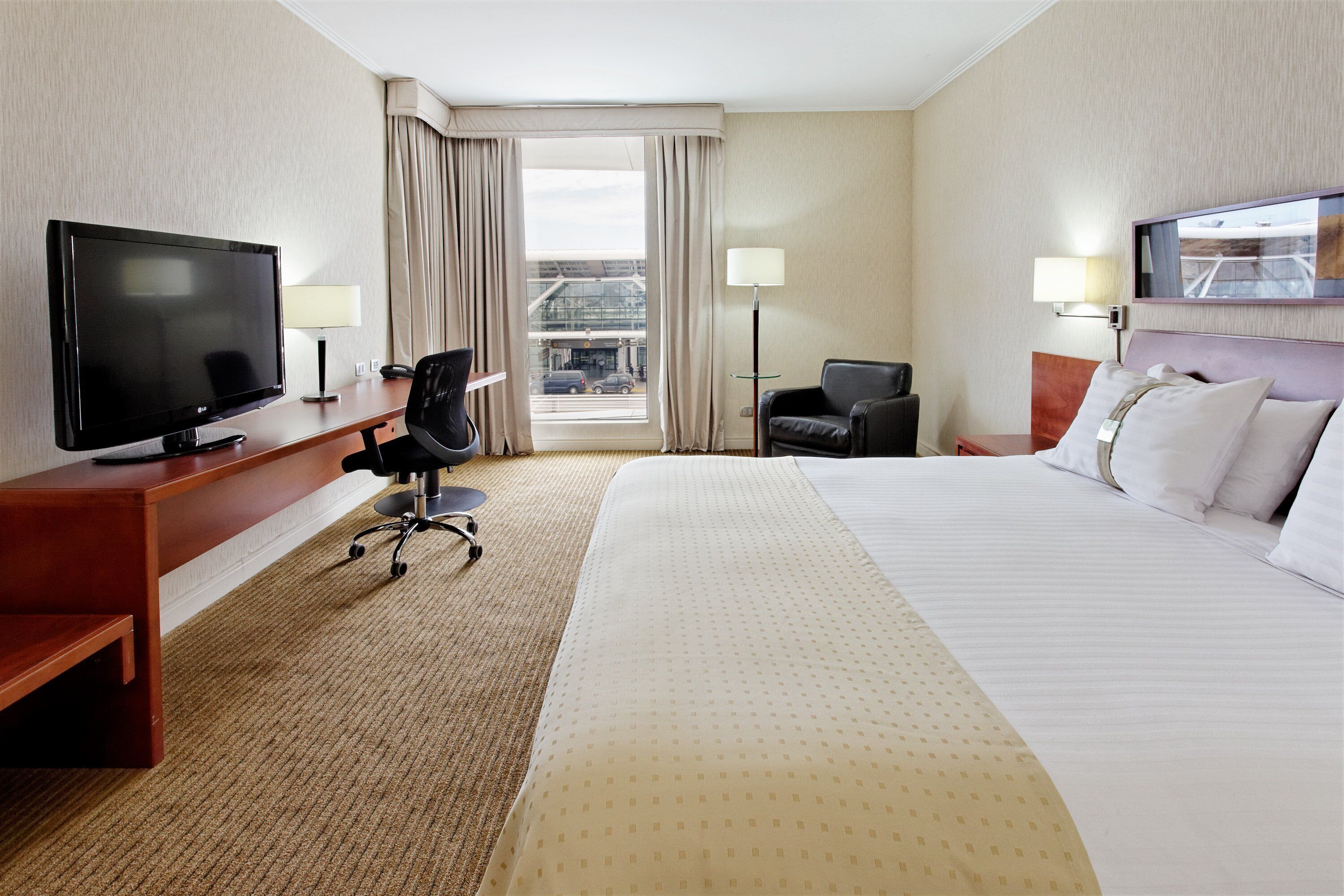 Quarto de hotel com uma cama, escrivaninha, televisão, cadeira, abajur e vista para o exterior.