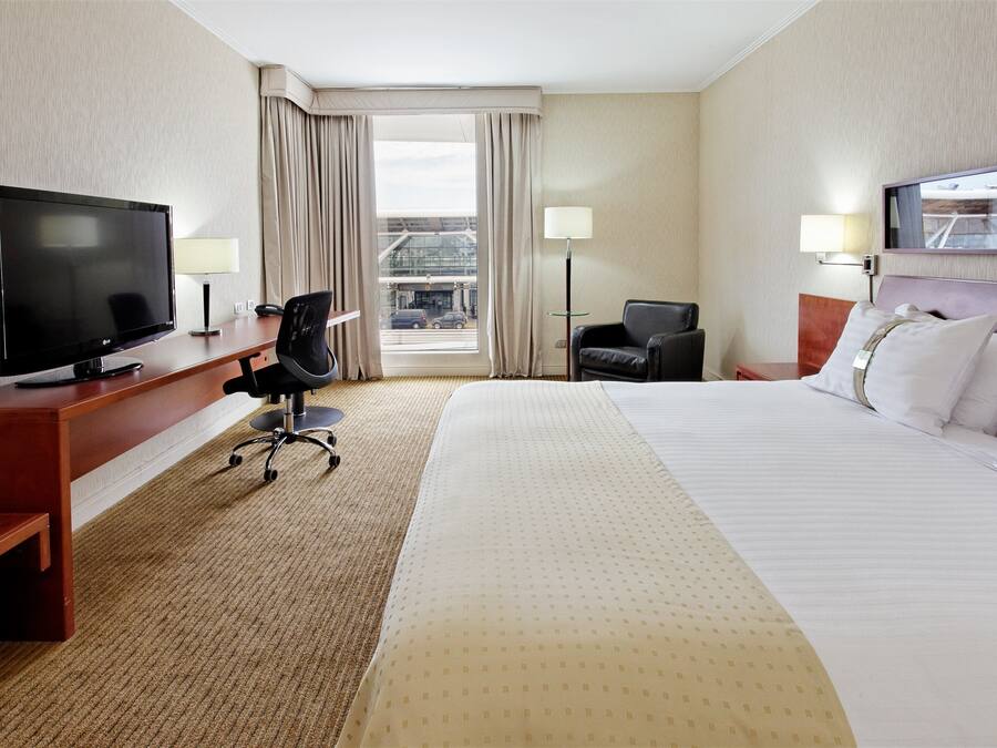 Quarto de hotel com uma cama, escrivaninha, televisão, cadeira, abajur e vista para o exterior.