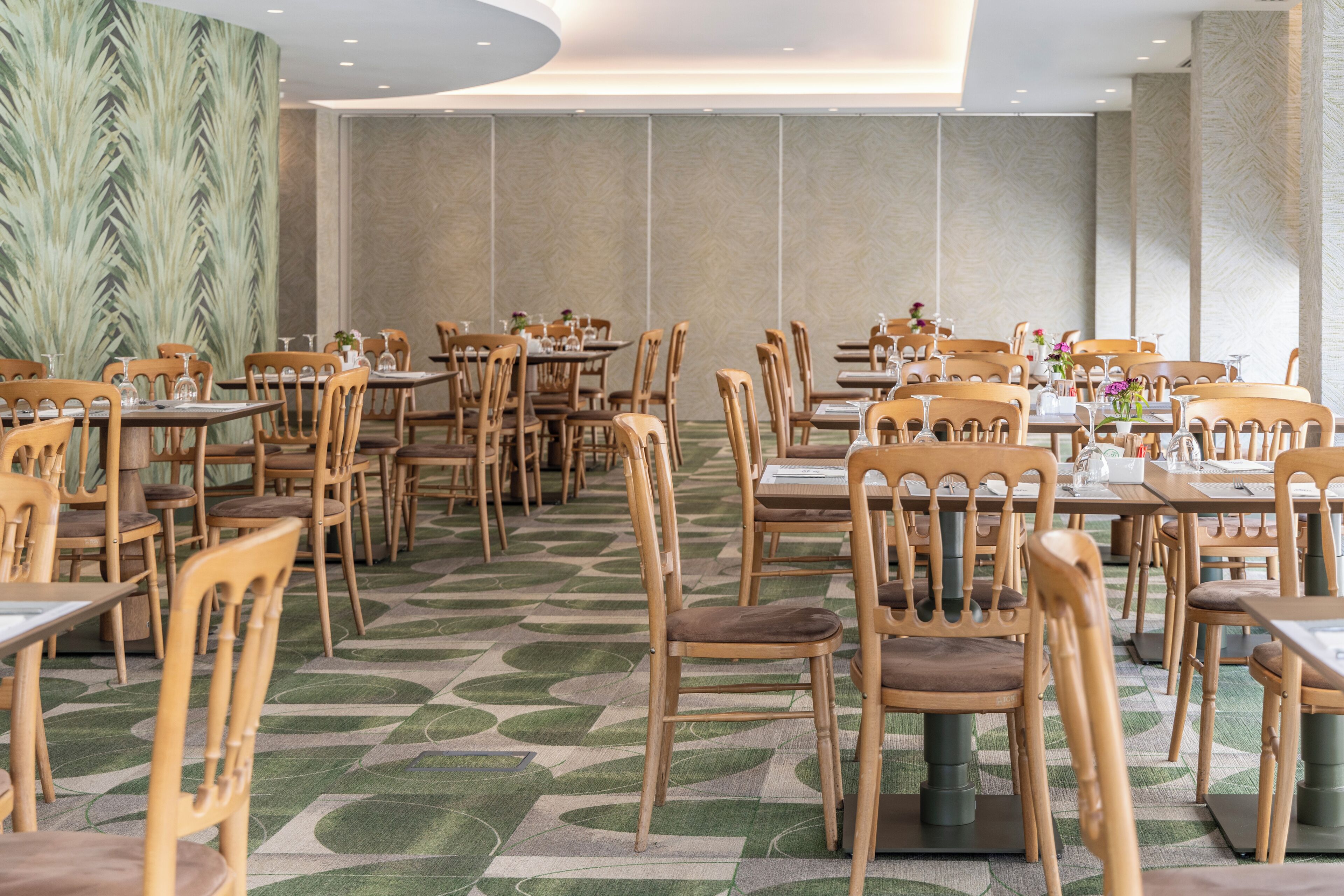 Um restaurante com cadeiras e mesas de madeira, um tapete estampado e uma parede verde com design de folhas.