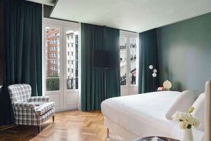 Quarto de hotel com uma cama grande, uma poltrona xadrez, uma televisão de tela plana e vista para a cidade através de grandes janelas.
