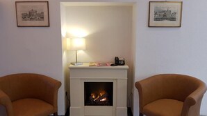 Um quarto com lareira, duas poltronas, um abajur e quadros nas paredes.