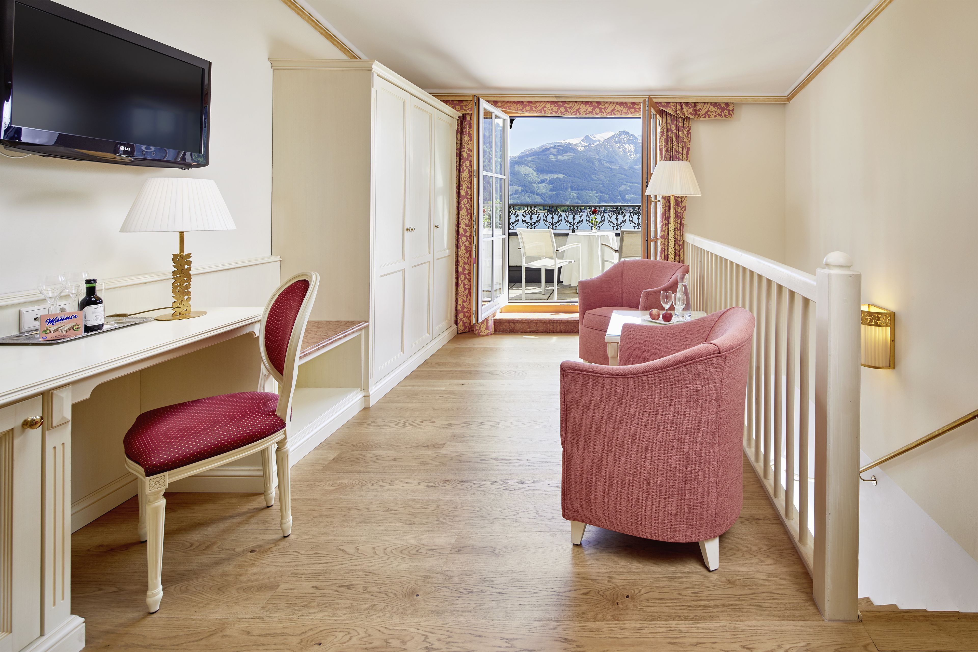 Quarto de hotel com piso de madeira, uma escrivaninha com abajur, televisão e duas cadeiras.