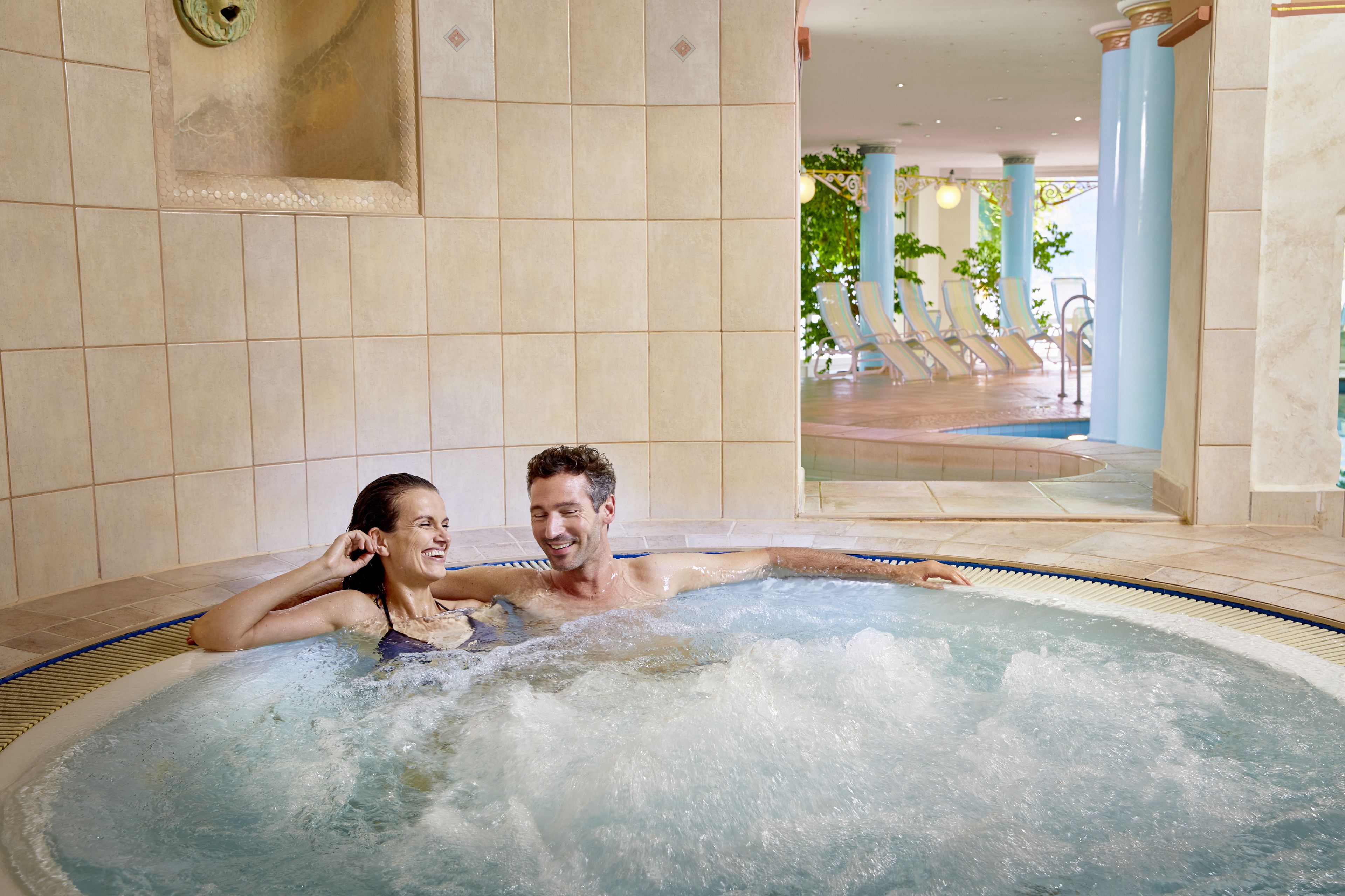 Um casal relaxando em uma banheira de hidromassagem com paredes e colunas azulejadas, com vista para uma área de piscina com espreguiçadeiras.