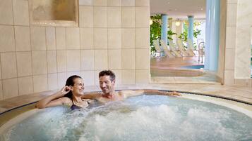 Um casal relaxando em uma banheira de hidromassagem com paredes e colunas azulejadas, com vista para uma área de piscina com espreguiçadeiras.
