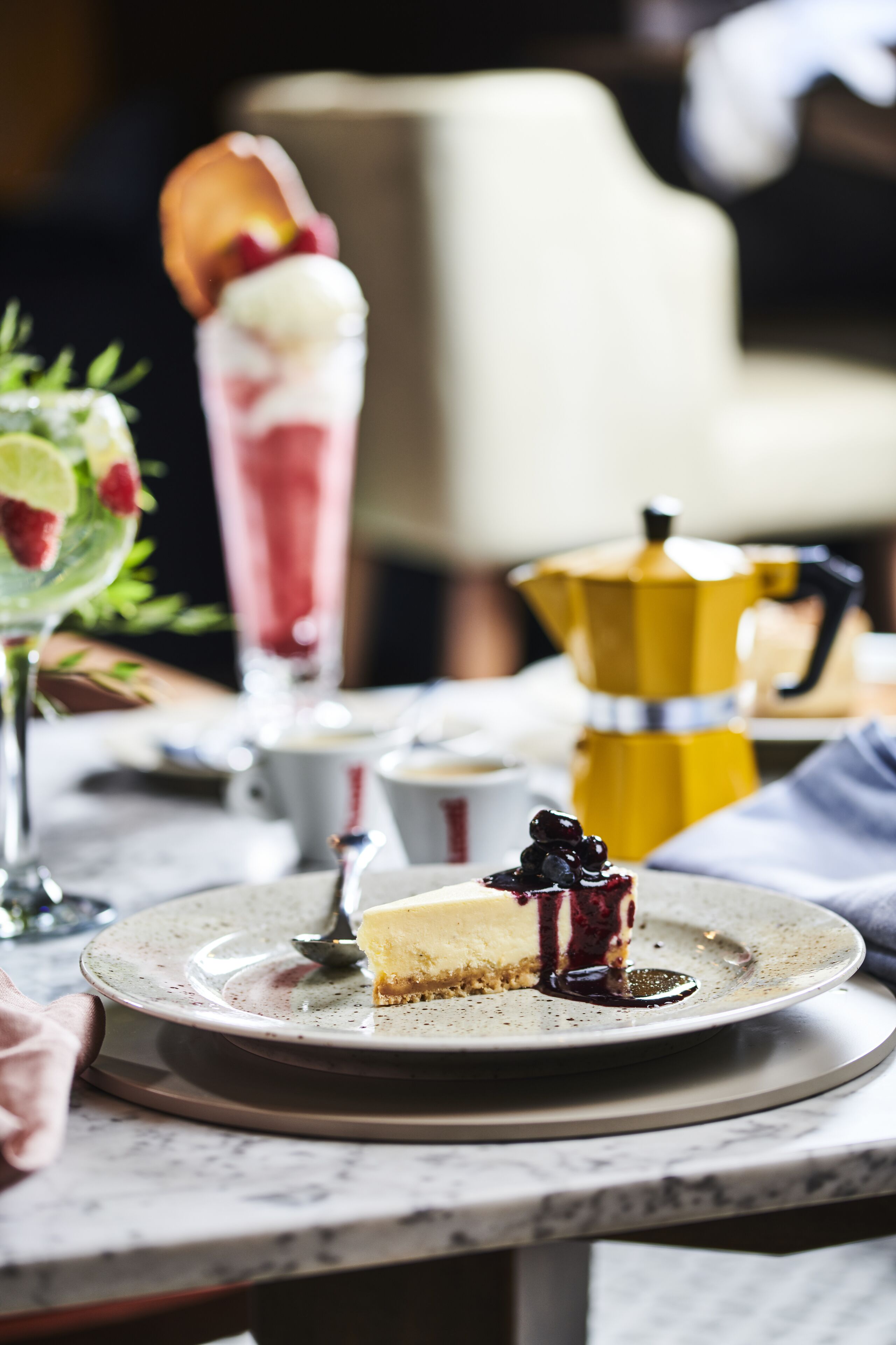Um prato com sobremesa, uma fatia de cheesecake e frutas vermelhas, uma bebida alta com guarnição de frutas, e uma cafeteira amarela ao fundo.