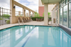 Piscina interior com água cristalina, cercada por espreguiçadeiras e mesas, amplas janelas e uma parede de tijolos.