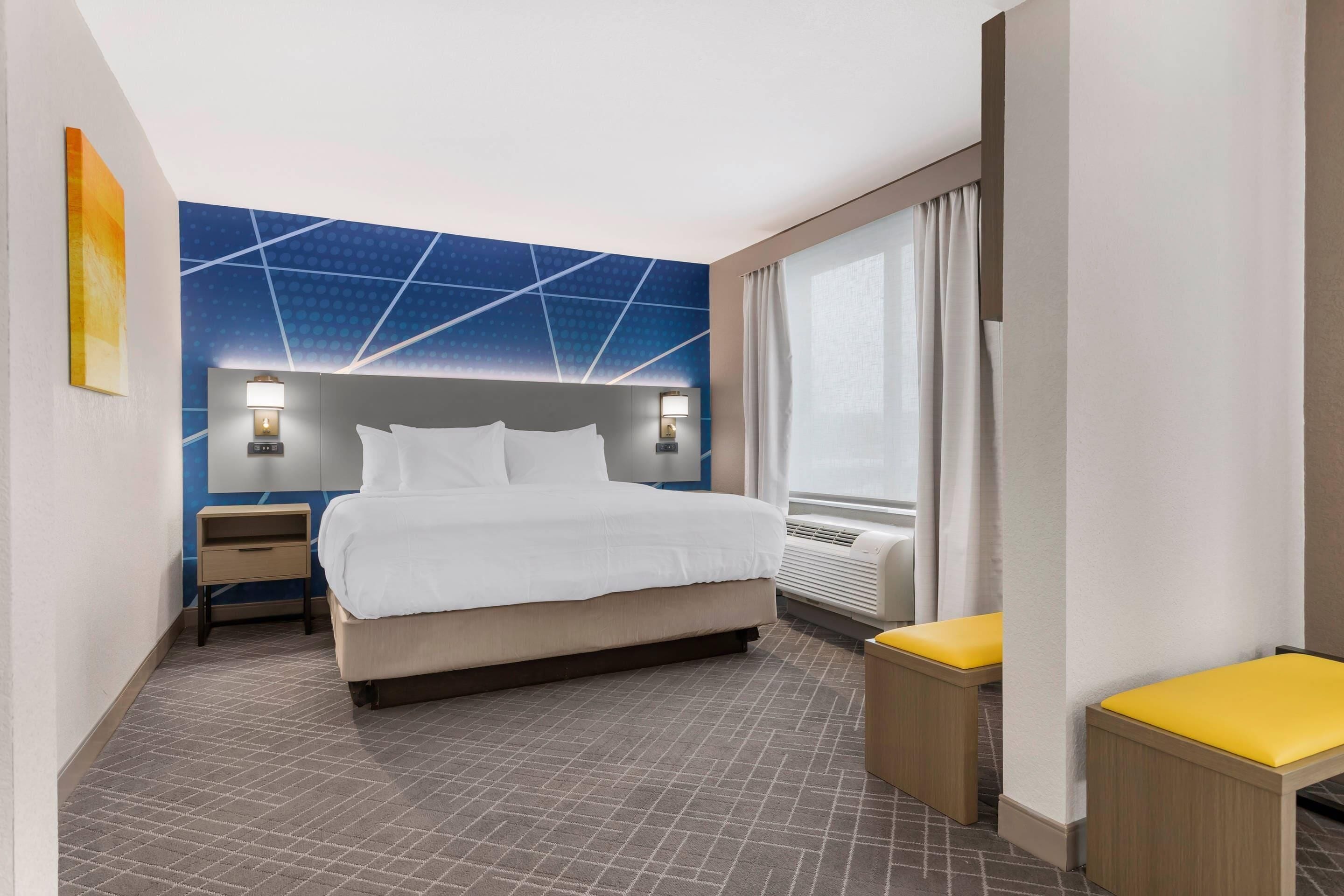 Quarto de hotel com cama, mesas de cabeceira, luminária na parede, janela com cortinas e piso estampado.