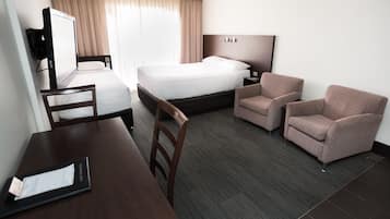 Quarto de hotel com uma cama, duas cadeiras, uma mesa de trabalho, televisão e janela com cortinas.